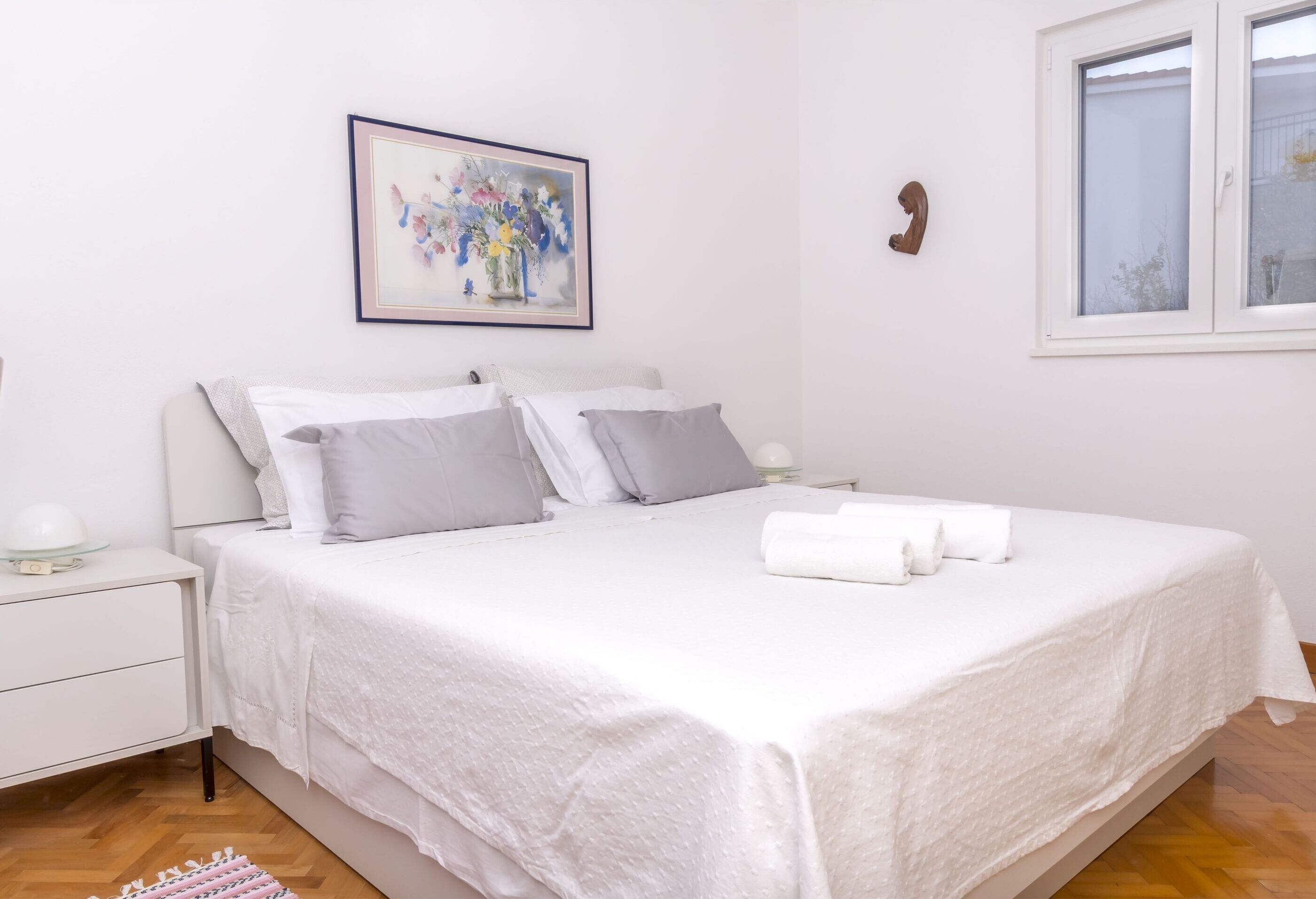Cama bem arrumada com lençóis brancos, mesa de cabeceira com abajur e um quadro com flores na parede.