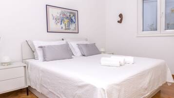 Cama bem arrumada com lençóis brancos, mesa de cabeceira com abajur e um quadro com flores na parede.