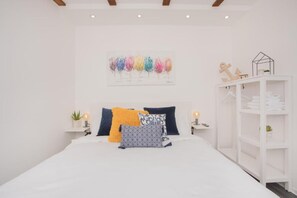 Um quarto bem arrumado com uma cama, almofadas, uma mesinha de cabeceira com abajur, uma obra de arte na parede e um móvel de estantes branco.