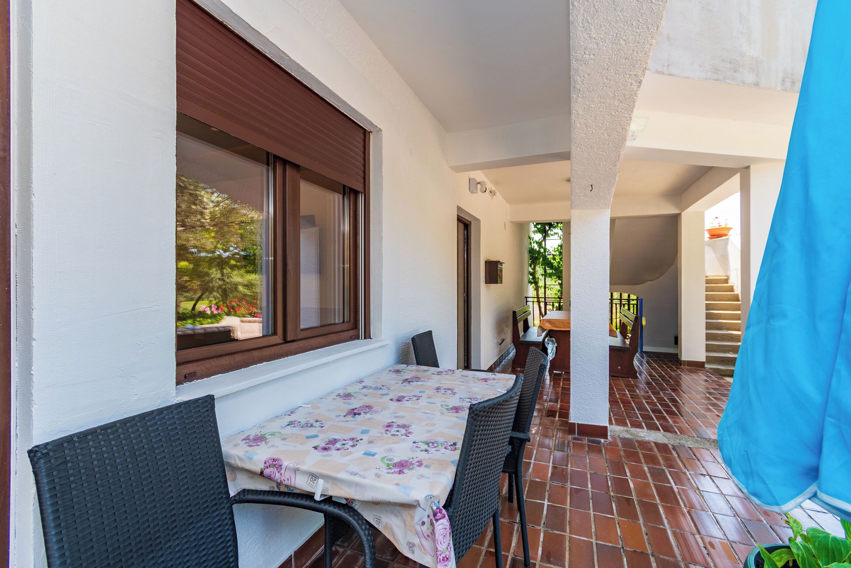 Um alpendre coberto com mesa e cadeiras, uma janela com persianas e uma escada que leva a um andar superior.
