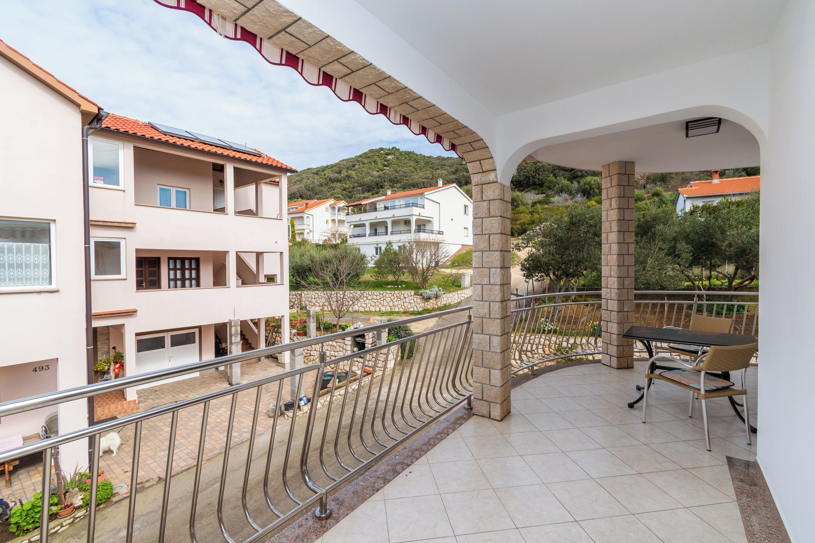 Varanda com guarda-corrimão de metal, uma mesa com cadeiras e vista para edifícios vizinhos e uma colina.