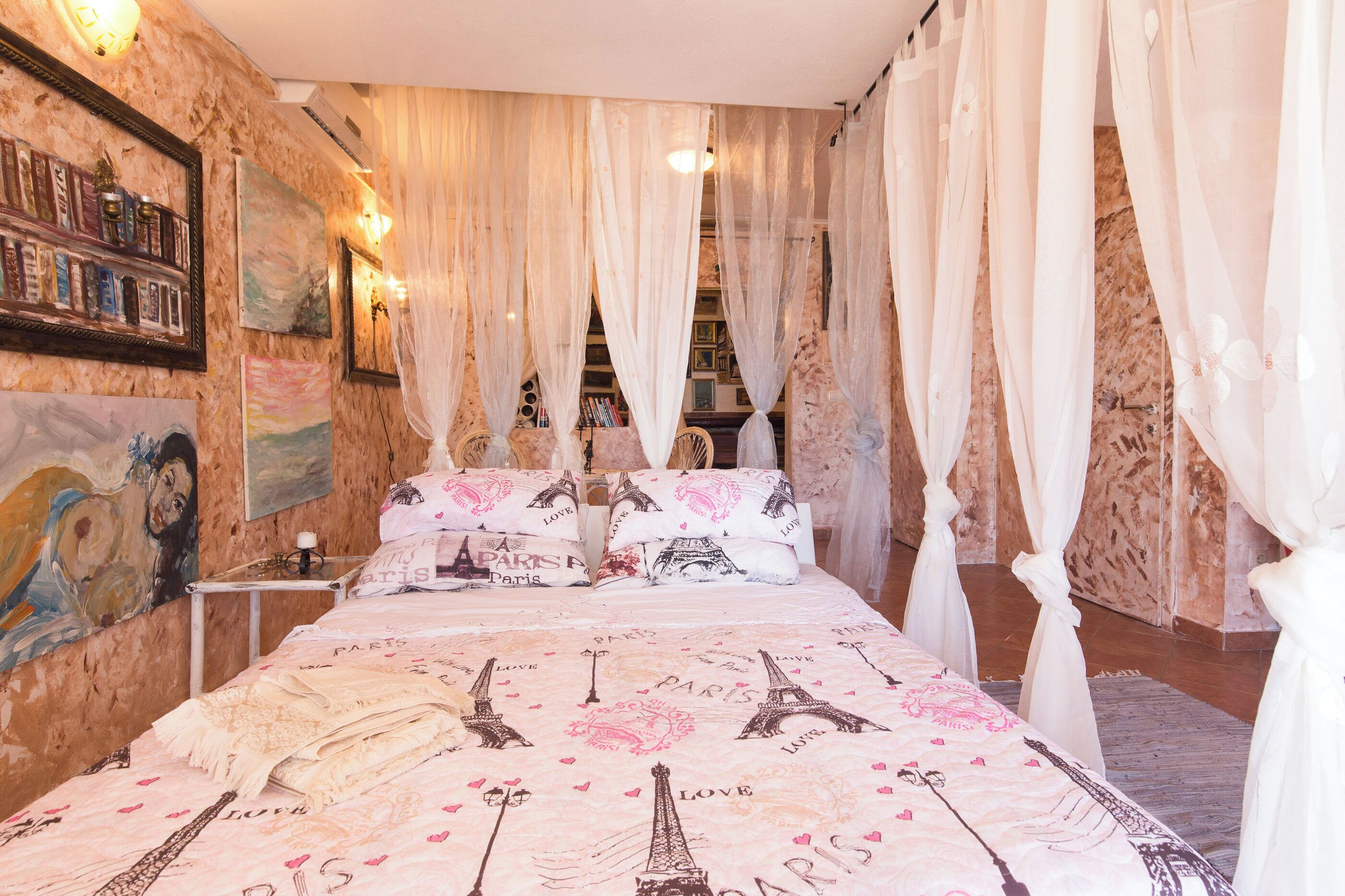 Quarto com cama decorada com roupa de cama com tema parisiense, uma estante de livros e um quadro na parede.