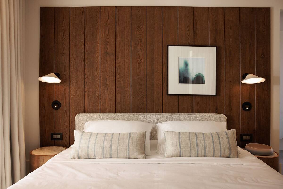 Uma cama de casal com duas almofadas, uma parede revestida de painéis de madeira, um quadro emoldurado e duas luminárias fixadas na parede.