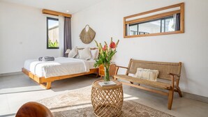 Quarto com cama, sofá de vime, mesa lateral redonda de madeira e espelho na parede.