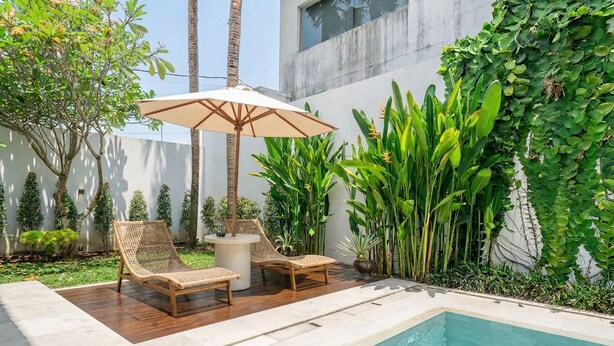 Uma área à beira da piscina com espreguiçadeiras, guarda-sol e vegetação exuberante.