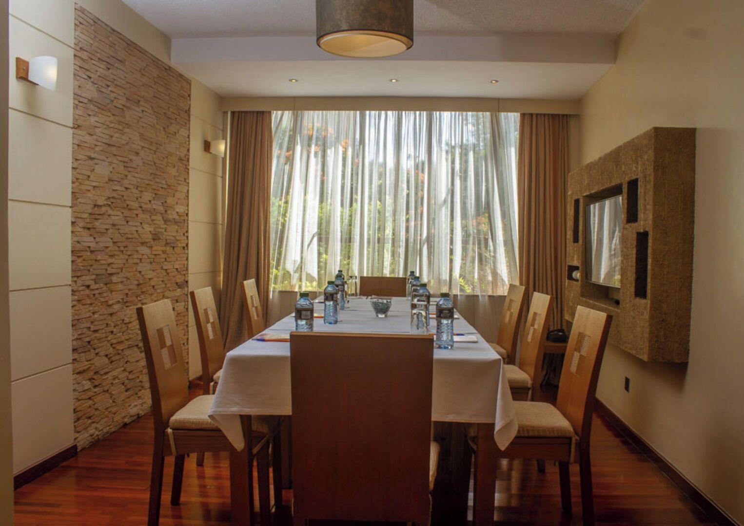 Uma sala de conferências com uma mesa comprida, cadeiras, garrafas de água e uma janela grande com cortinas.