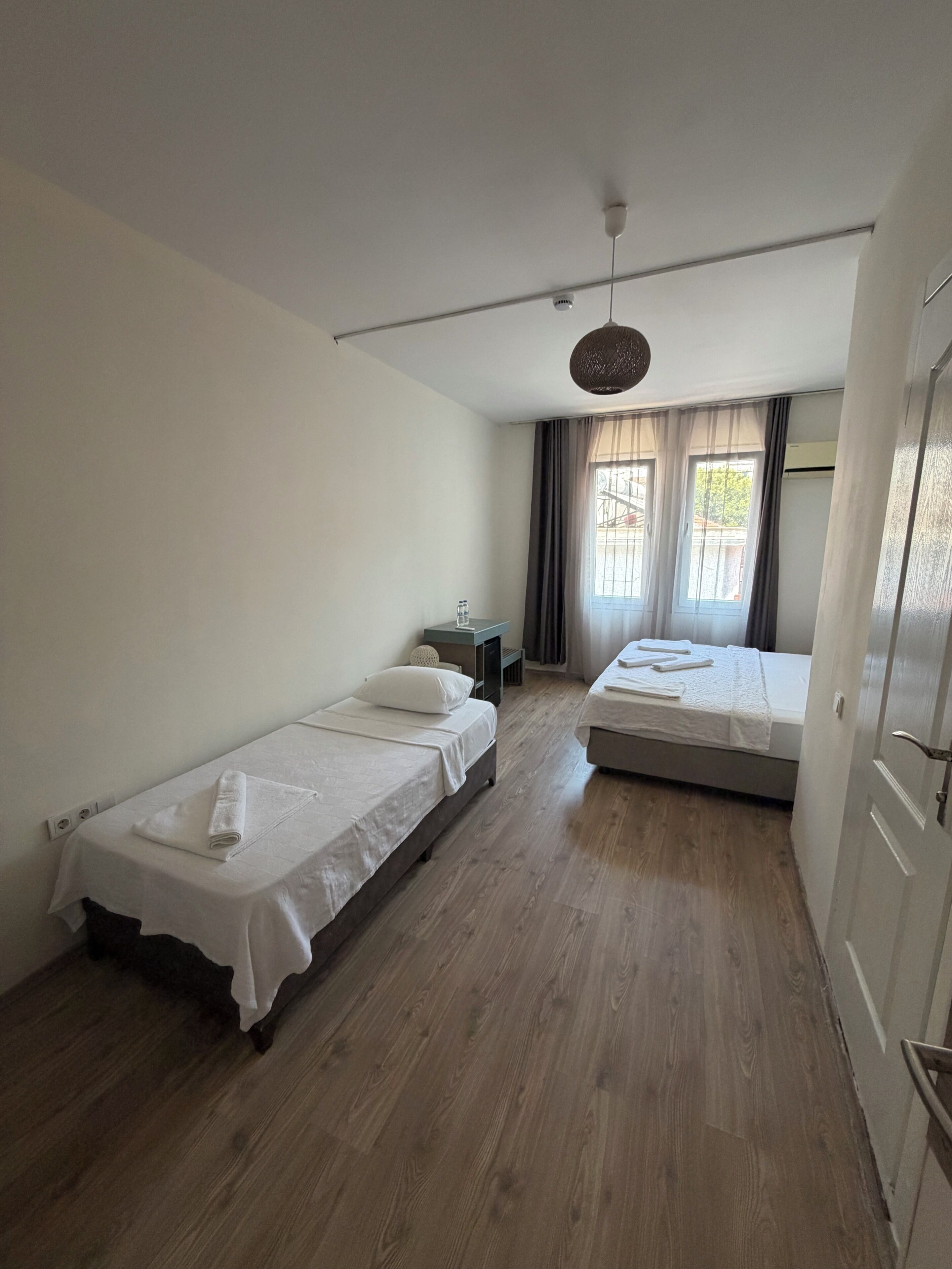 Quarto com duas camas de solteiro, uma mesa pequena, um lustre e uma porta.
