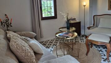 Uma sala de estar com um sofá, uma poltrona, uma mesinha com livros, um abajur e uma janela com cortinas.