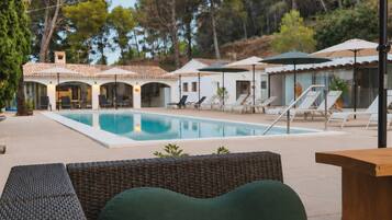Uma área de piscina com espreguiçadeiras e guarda-sóis, rodeada por árvores e edifícios.