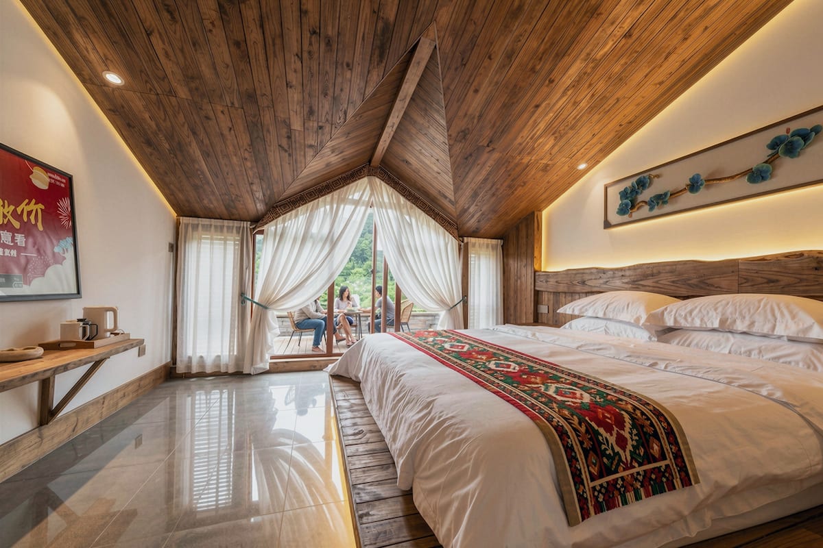 Quarto com teto de madeira, cama com roupa de cama branca, um painel decorativo na parede e vista pela janela com cortinas leves.