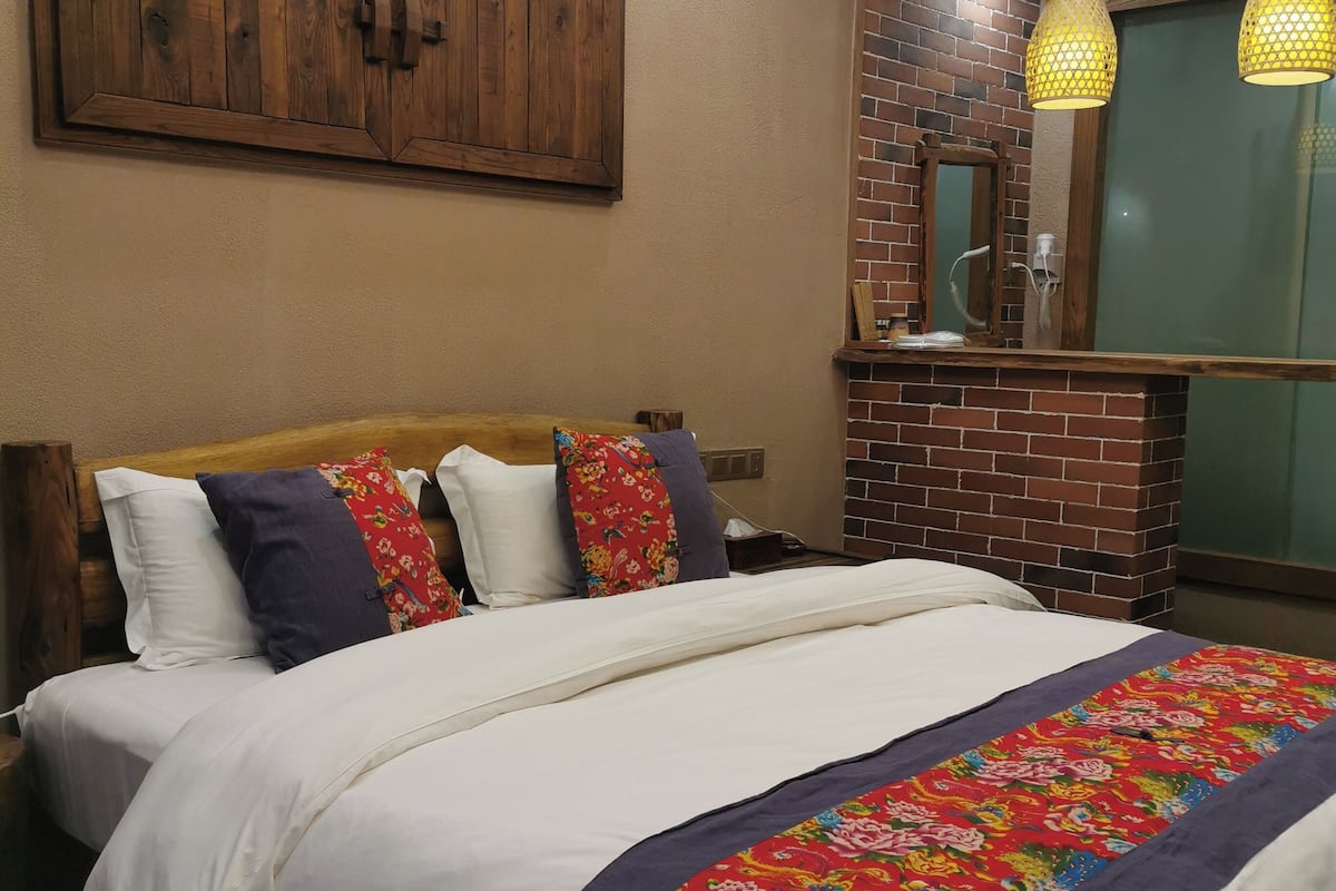 Quarto com cabeceira de madeira, cama com roupa de cama branca e manta floral, parede de tijolos e luminária pendente.
