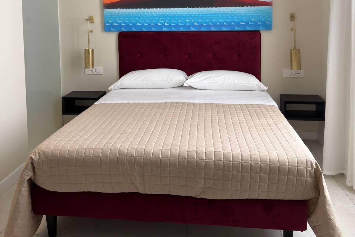 Uma cama com cabeceira vermelha, um quadro de um vulcão e oceano, e abajures nas mesas de cabeceira.