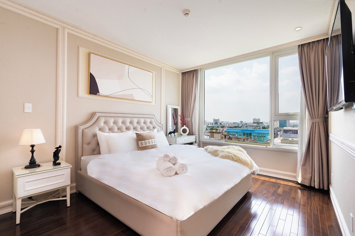 Quarto de hotel com uma cama grande, mesa de cabeceira com abajur, televisão e vista para a cidade.