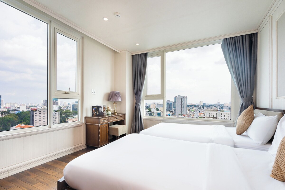Quarto de hotel com duas camas, uma escrivaninha e grandes janelas com vista para a cidade.