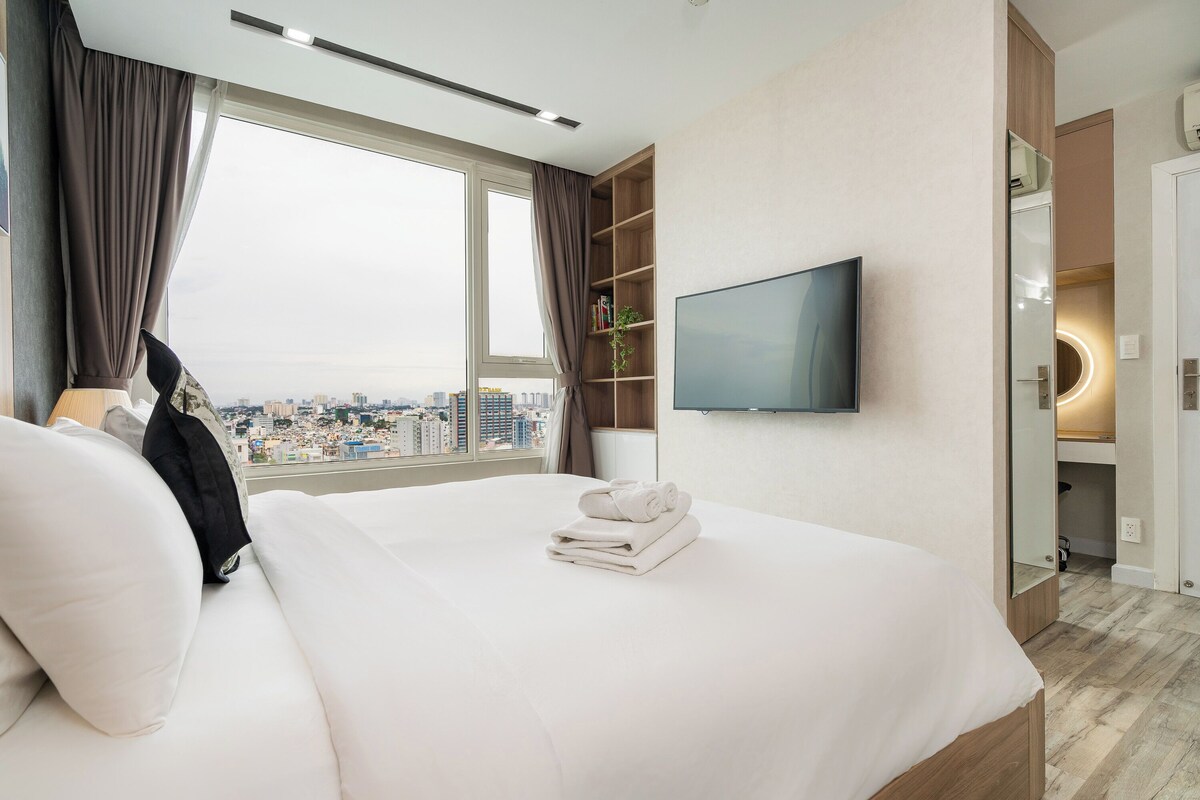Quarto de hotel com uma cama grande, televisão fixada na parede, vista para a cidade e uma estante de madeira.