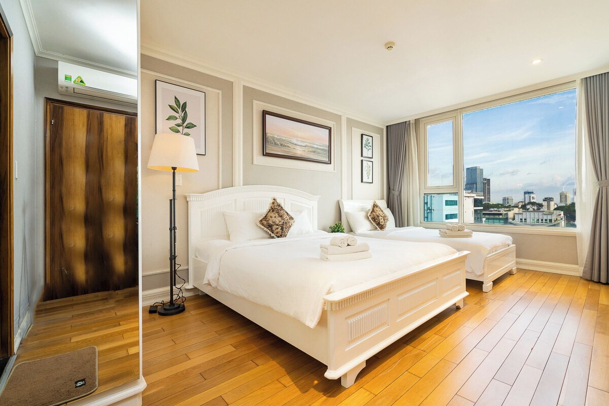 Quarto de hotel com uma cama grande, uma luminária de chão, vista para a cidade e uma janela com cortinas.