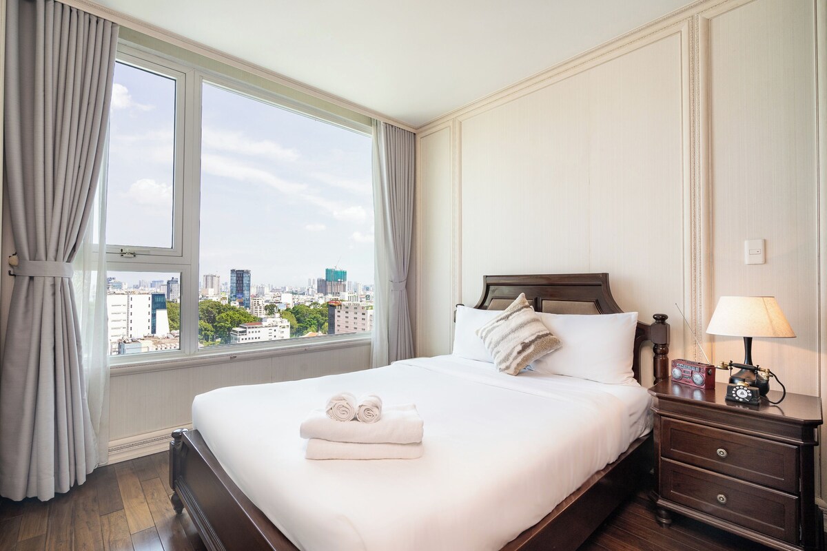 Quarto de hotel com uma janela grande, cama bem arrumada, mesa de cabeceira com abajur e vista para a cidade.