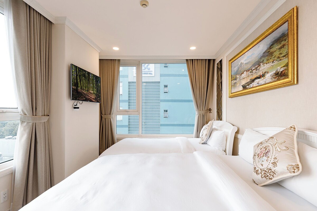 Quarto de hotel com uma cama grande, um quadro, vista de um edifício e cortinas.