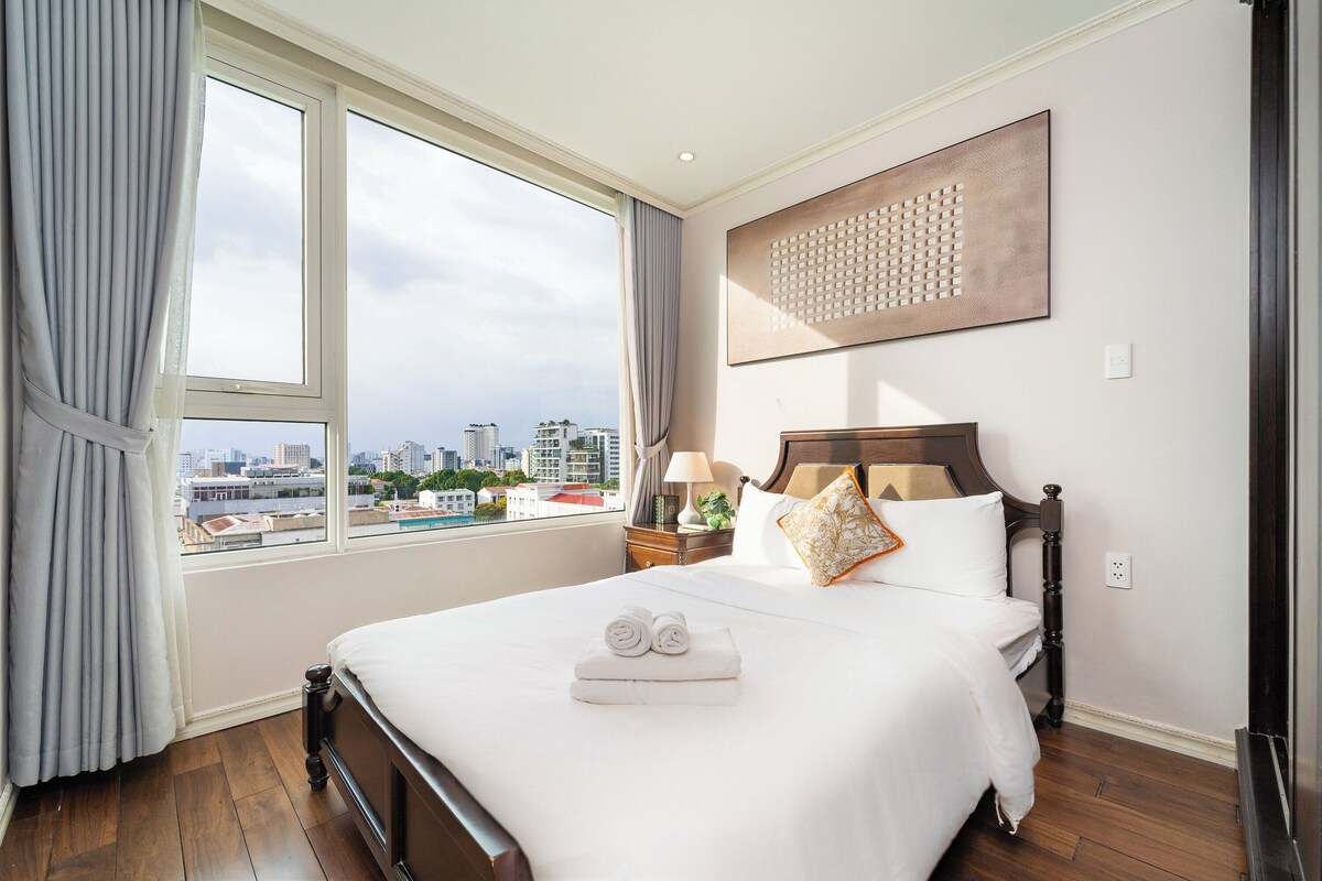 Quarto de hotel com uma janela grande, cama com lençóis brancos, mesa de cabeceira com abajur e vista para a cidade.