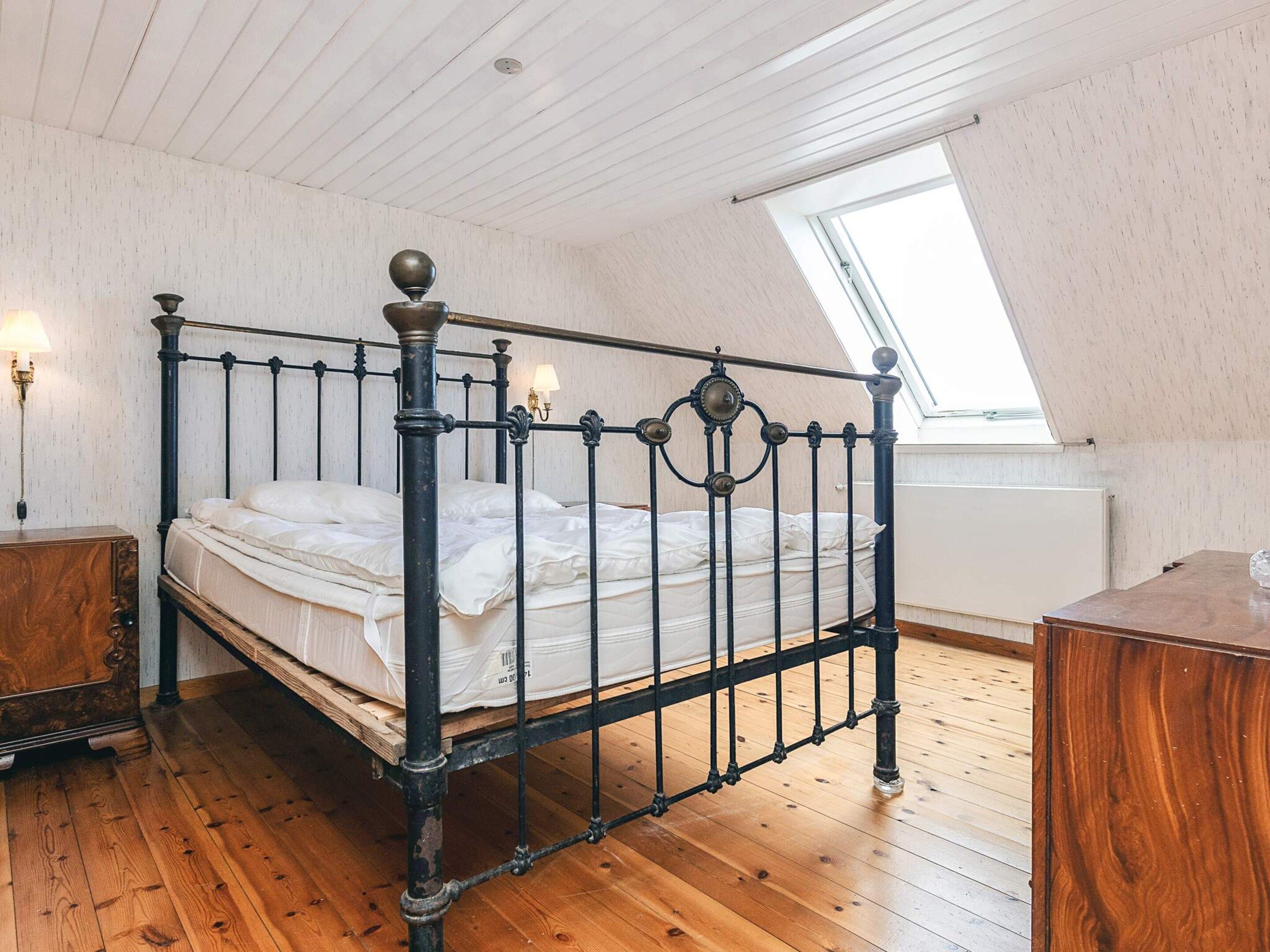 Quarto com estrutura de cama em metal, piso de madeira, claraboia e mesa de cabeceira com abajur.