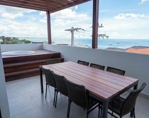 Um alpendre coberto com mesa e cadeiras de madeira, um espaço de estar pequeno com degraus e vista para o mar.