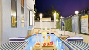 Uma área de piscina com espreguiçadeiras, mesa com petiscos e bebidas, e uma placa com as regras da piscina.