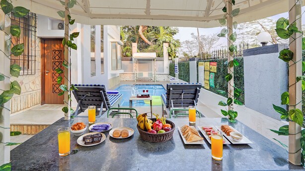Café da manhã à beira da piscina, com cadeiras, mesa posta com comida e bebidas, e vista para a piscina e vegetação.