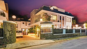 Um edifício moderno de dois andares com entrada gradeada, uma placa com o nome 'ADARSH' e um carro estacionado na frente.