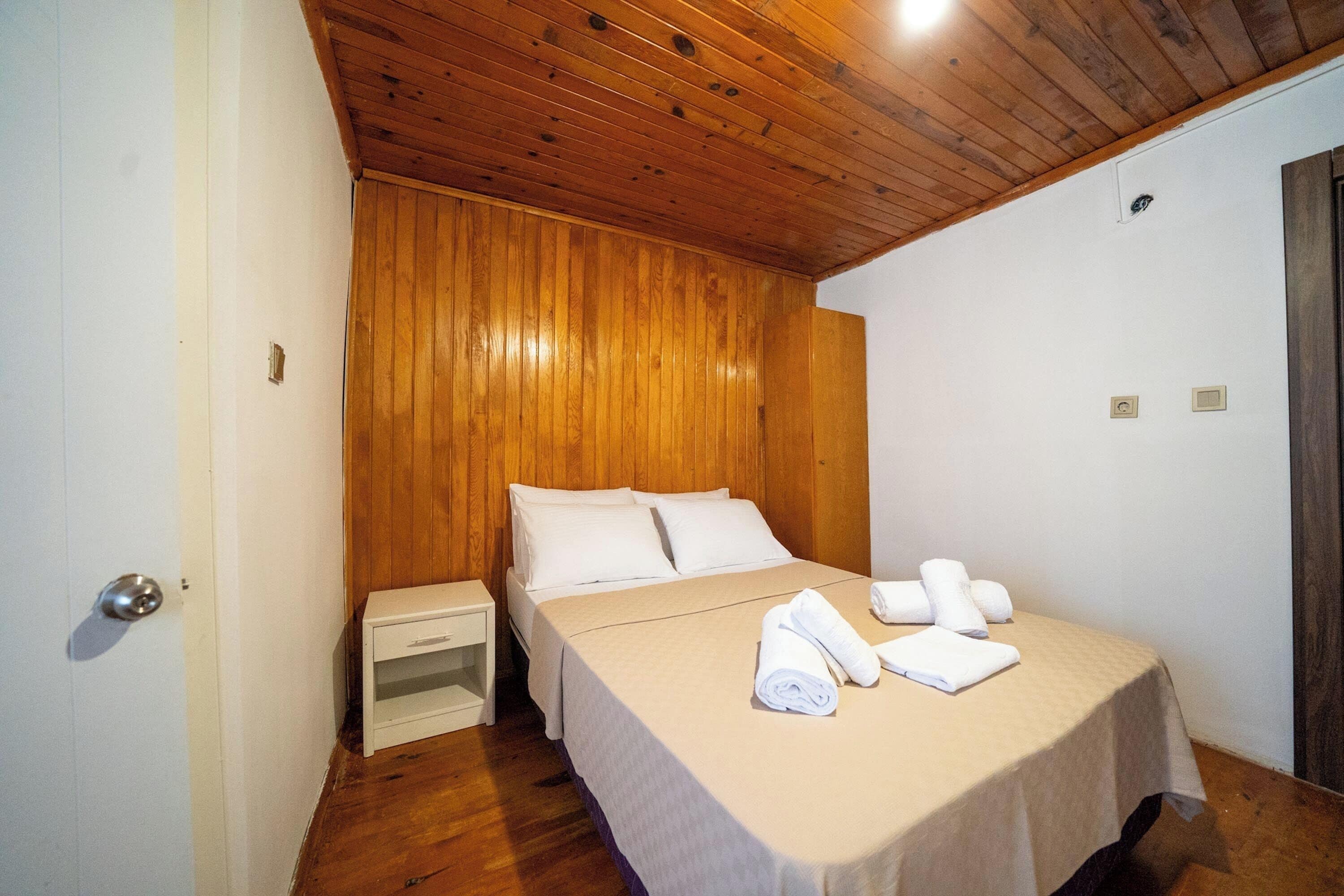 Uma cama de solteiro com roupa de cama e toalhas brancas, teto de madeira, uma pequena mesa de cabeceira e uma parede branca.