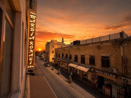 Vista da rua ao pôr do sol, com edifícios, uma placa indicando 'Mardi Gras' e uma loja chamada 'BIFE KELES'.