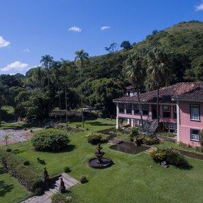Um edifício rosa com telhado ladrilhado, cercado por um jardim bem cuidado com uma fonte, palmeiras e um lago.
