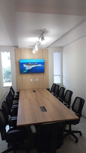 Uma sala de conferências com uma mesa comprida, cadeiras de escritório pretas, uma televisão instalada e um lustre no teto.