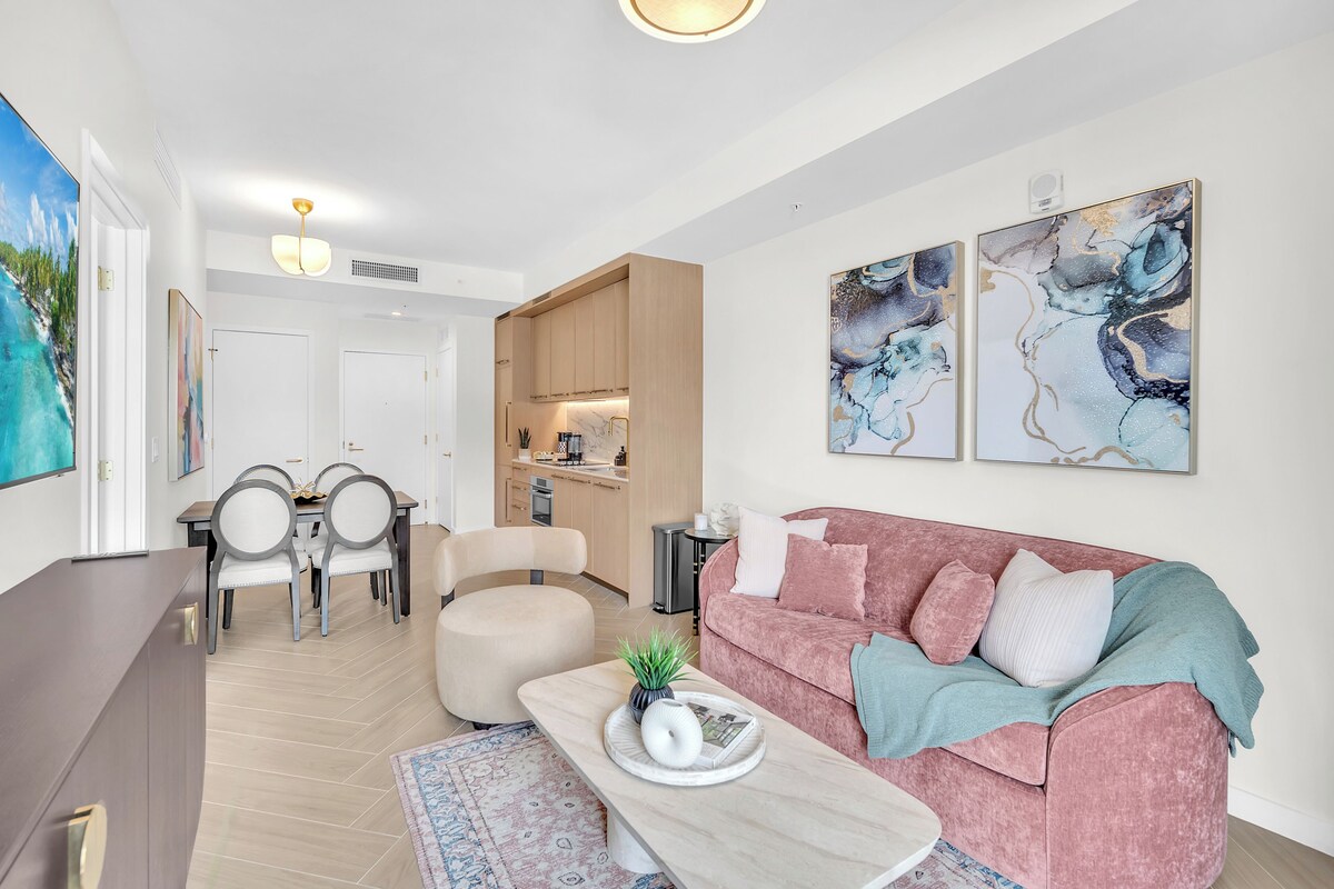Uma sala de estar moderna com um sofá rosa, uma área de jantar com mesa e cadeiras, e uma cozinha ao fundo.