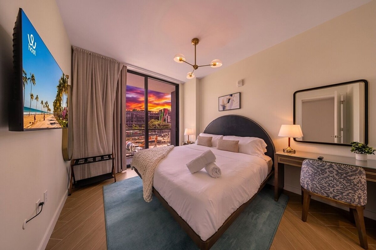 Quarto de hotel com uma cama grande, uma escrivaninha, uma cadeira, televisão e vista para um pôr do sol e paisagem urbana.