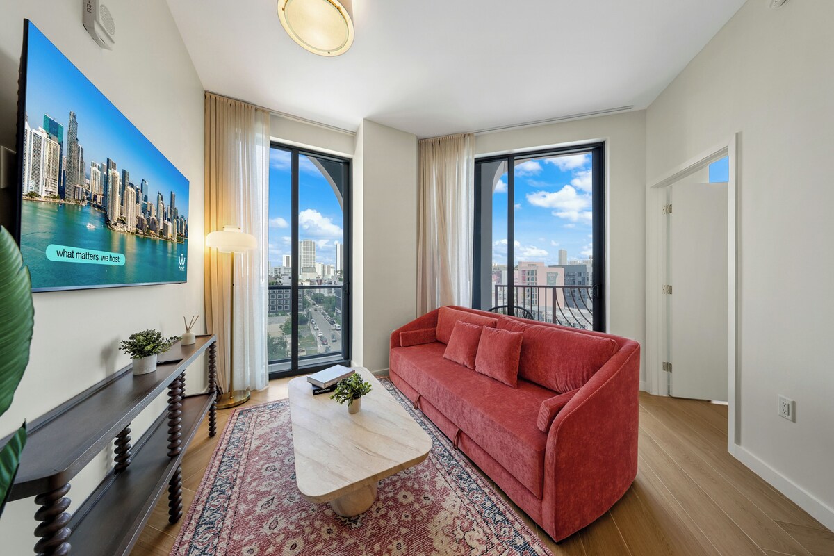 Uma sala de estar moderna com um sofá vermelho, uma mesa de centro, uma grande janela com vista para a cidade e um quadro na parede.