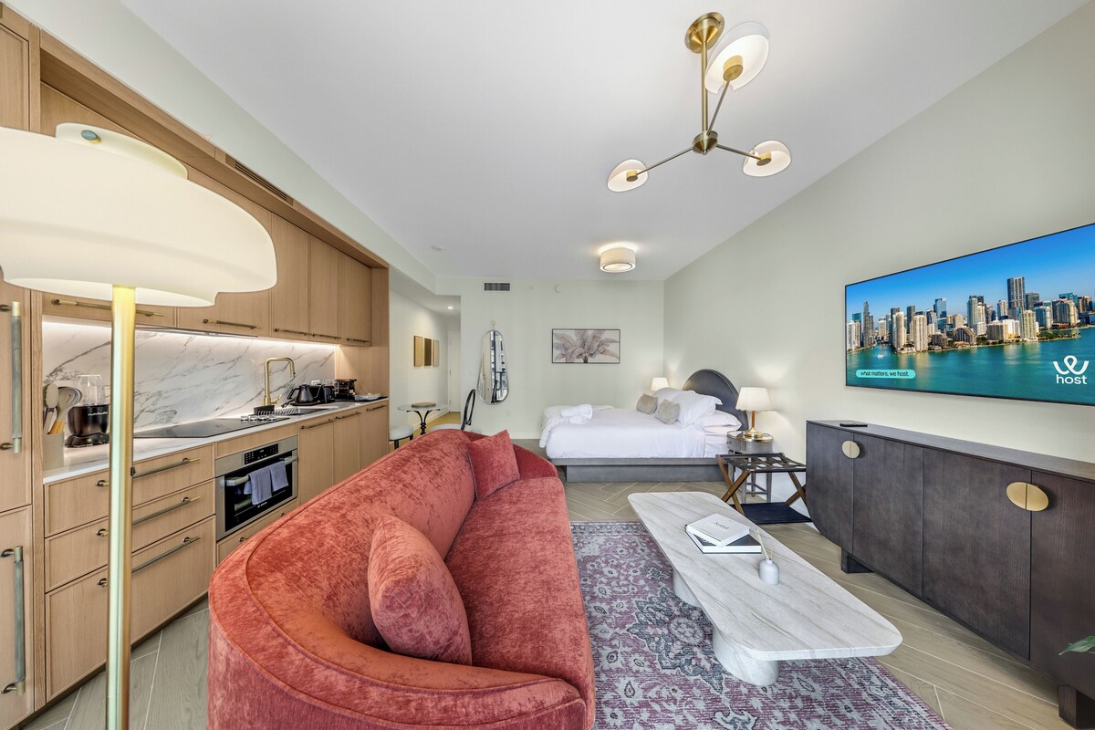 Quarto de hotel moderno com cozinha, um sofá vermelho, uma TV exibindo uma vista urbana e uma cama.