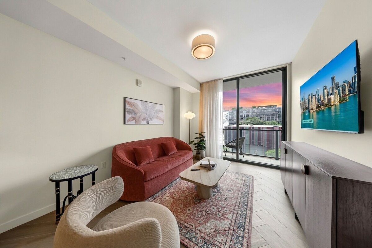 Uma sala de estar moderna com um sofá vermelho, uma mesa de centro, uma televisão na parede e uma janela grande com vista para a cidade.