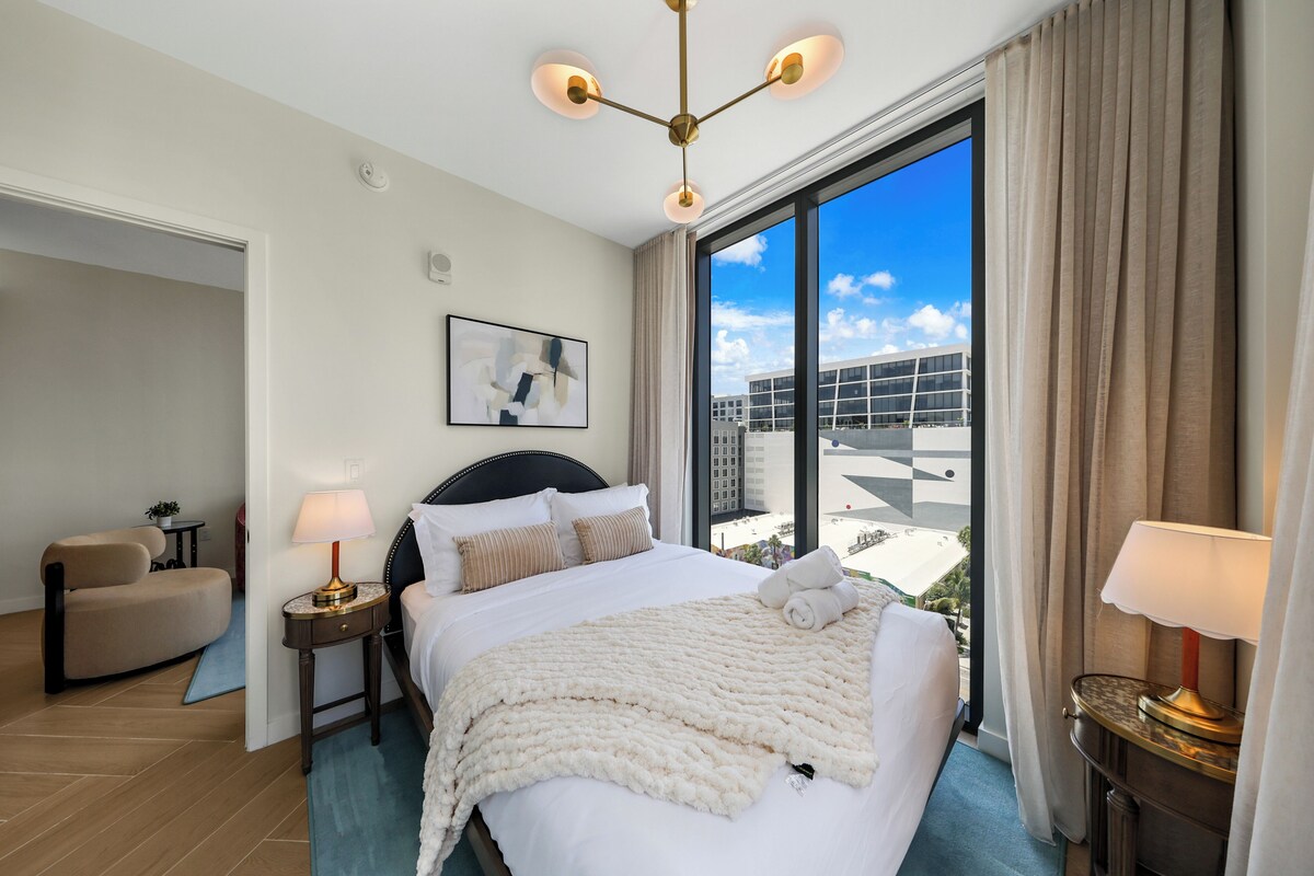 Quarto de hotel com uma cama grande, uma mesinha de cabeceira com abajur, uma cadeira, uma janela com vista para edifícios e um lustre.