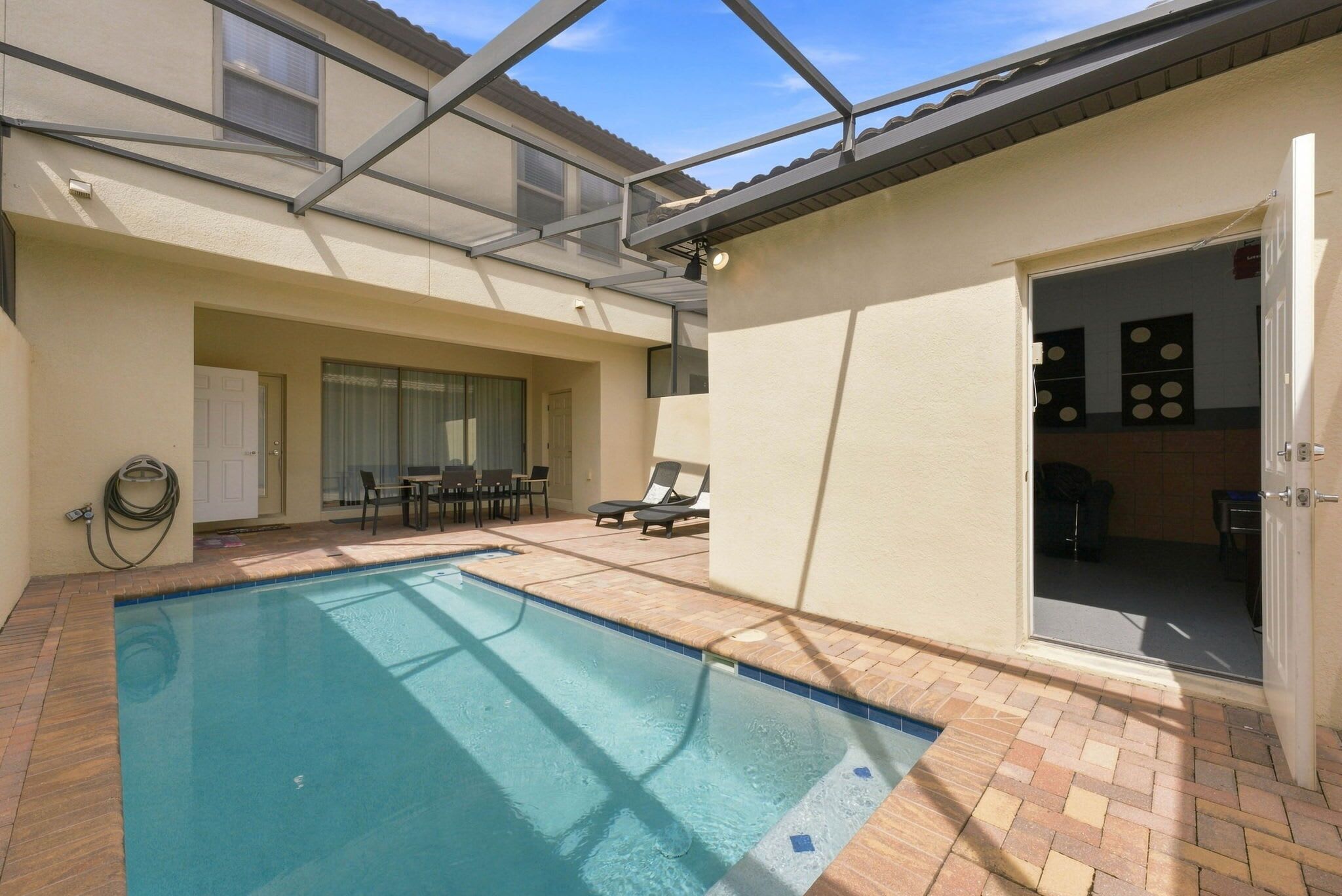 Uma piscina com área de terraço, um espaço de estar coberto e uma porta que dá para um quarto com piso ladrilhado.