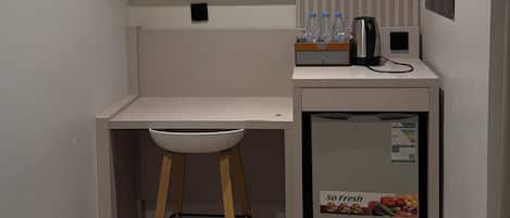 Quarto de hotel compacto com escrivaninha, pequeno frigorífico, um banquinho, dispensador de água e janela com persianas.