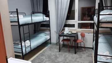 Um dormitório com beliches, uma pequena mesa com cadeiras e uma janela com cortinas.