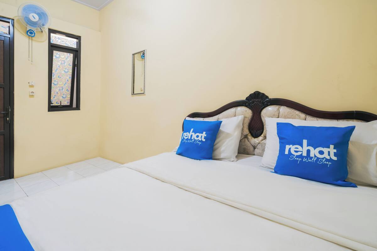Quarto de hotel com cama, duas almofadas azuis com a marca 'rehat', janela com cortinas estampadas e ventoinha fixada na parede.