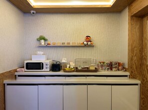 Uma cozinha compacta com micro-ondas, torradeira e uma pequena geladeira. Há prateleiras acima com diversos objetos e uma câmera na parede.