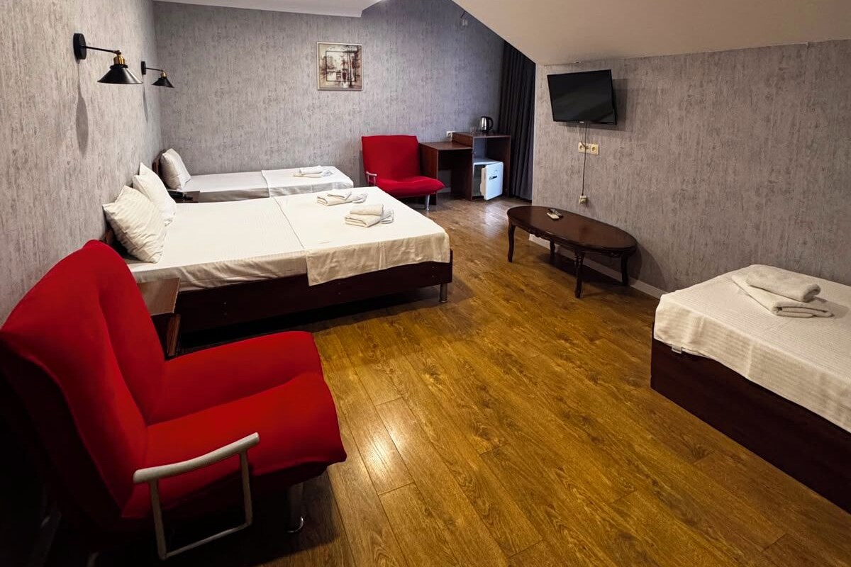 Quarto de hotel com duas camas, uma poltrona vermelha, piso de madeira, televisão de tela plana e um quadro na parede.
