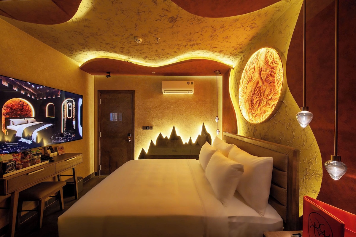 Quarto de hotel com cama, escrivaninha, televisão e ar-condicionado fixado na parede.