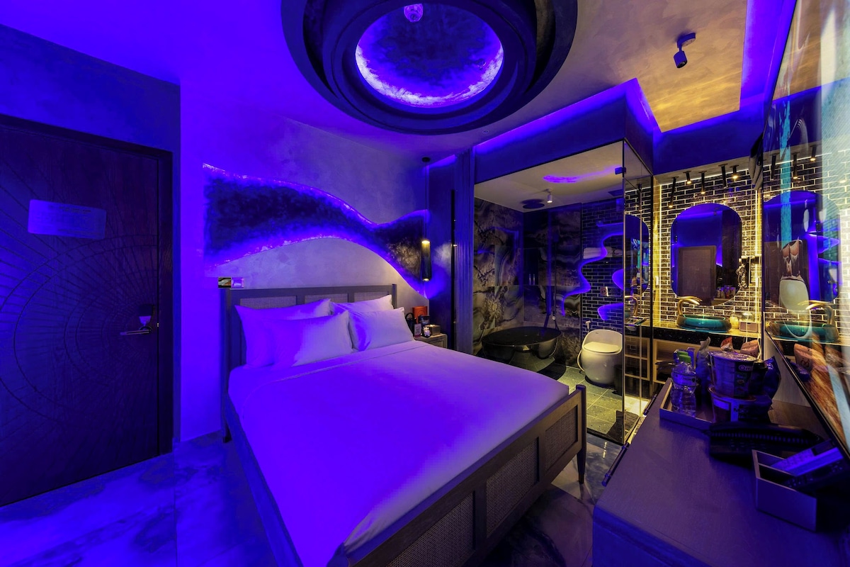 Quarto de hotel moderno com cama, luminária redonda no teto, banheiro com espelho e parede com design texturizado.