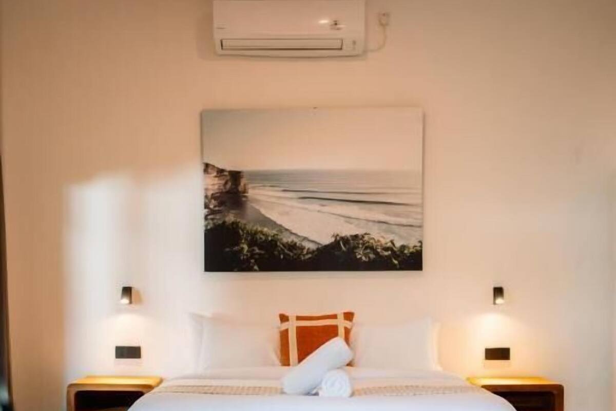Quarto com cama, um banquinho, duas mesas de cabeceira, ar-condicionado fixado na parede e um quadro.