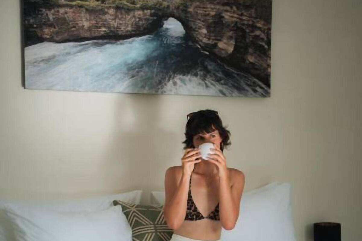 Uma cama com roupa de cama branca, um travesseiro com padrão geométrico e uma pintura de paisagem acima da cama.