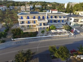 Um edifício de vários andares com varandas, uma área pavimentada com mesas ao ar livre e uma rua em primeiro plano.