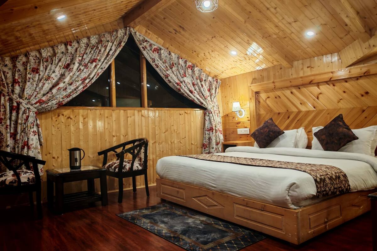 Quarto com painéis de madeira, uma cama, uma mesinha de cabeceira, uma cadeira e uma janela com cortinas.